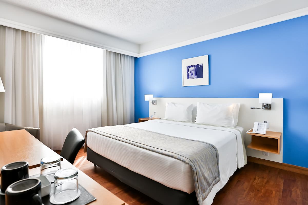 Quarto de hotel com uma cama grande, uma mesa de madeira com xícaras de café, um abajur e um telefone em uma mesa de cabeceira.