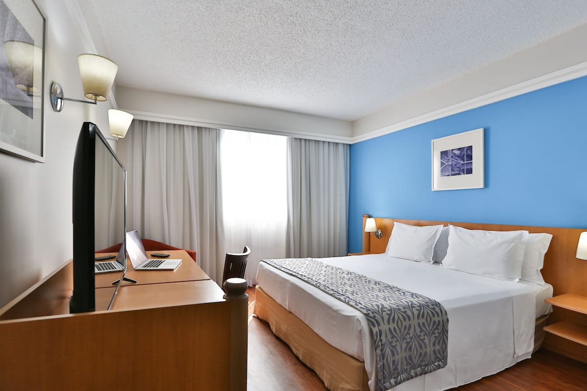 Quarto de hotel com cama, escrivaninha com um laptop, cadeira, mesa de cabeceira com abajur e janela com cortinas.
