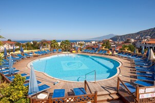 Uma área de piscina com cadeiras de descanso azuis e guarda-sóis, cercada por um deck de madeira e palmeiras.