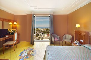 Quarto de hotel com varanda, uma cama, escrivaninha com TV, cadeira e telefone.