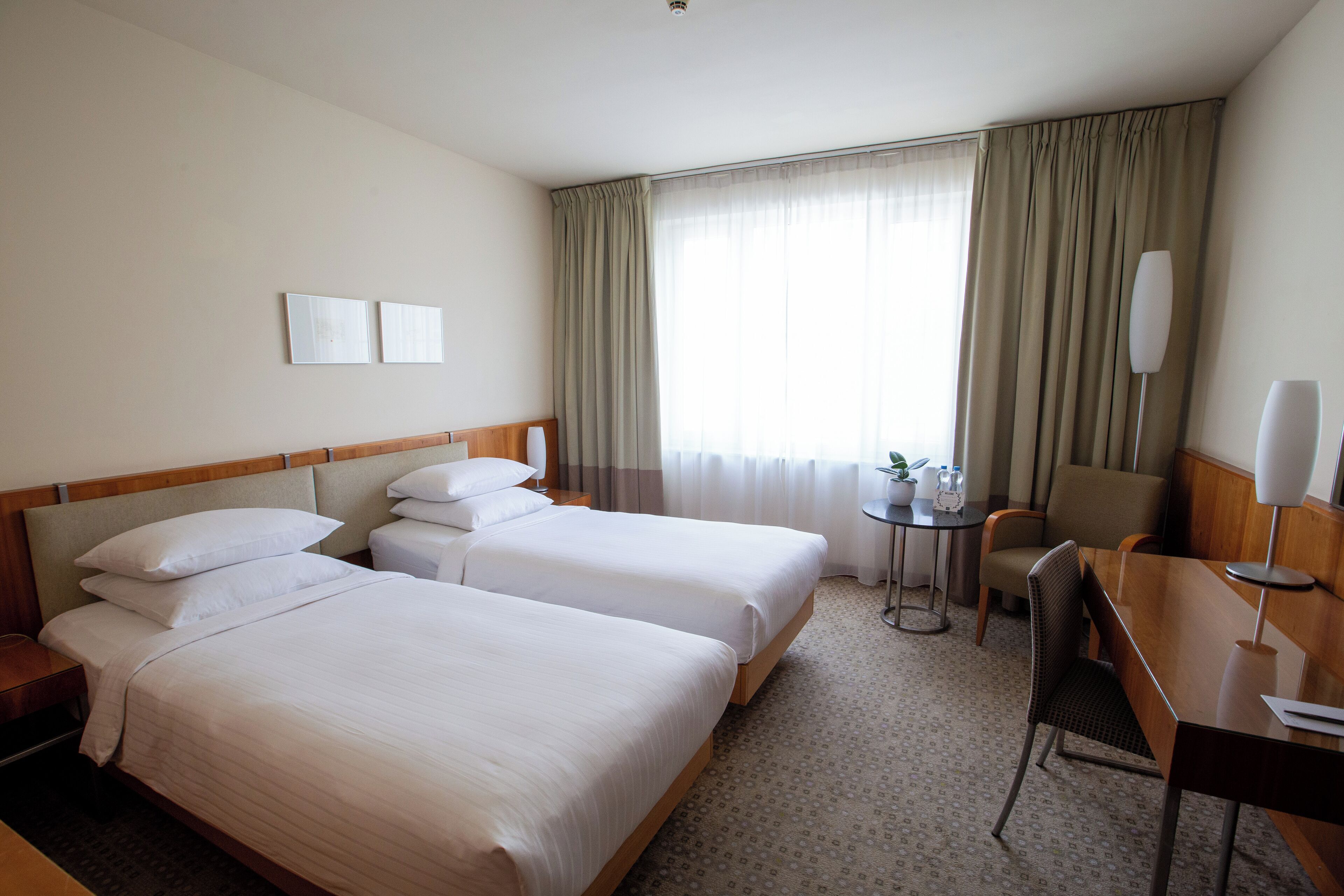 Quarto de hotel com duas camas de solteiro, uma pequena mesa de escritório com cadeira, uma mesinha de cabeceira e uma janela com cortinas.