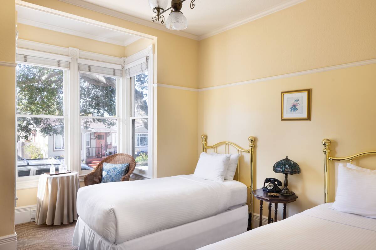 Quarto de hotel com duas camas, janela com vista para o exterior, uma mesinha com abajur e um quadro pendurado na parede.