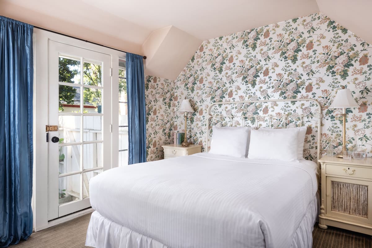Quarto com uma cama grande, papel de parede floral, mesa de cabeceira com abajur e porta com cortinas azuis.