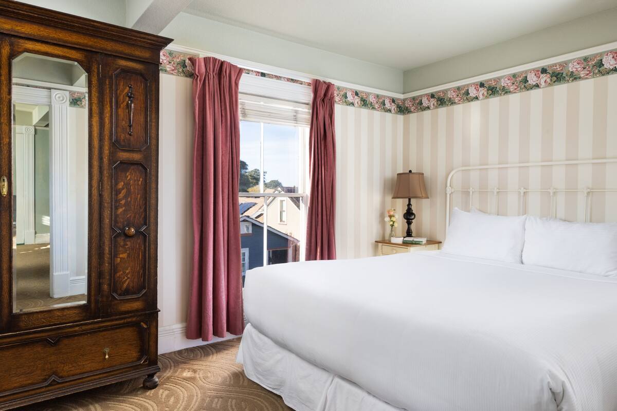 Quarto de hotel com uma cama grande, cômoda de madeira, mesa de cabeceira com abajur e janela com cortinas.