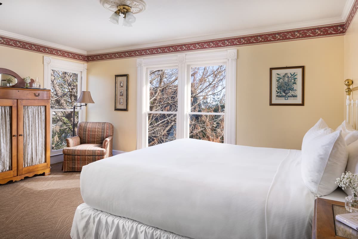 Quarto com uma cama grande, penteadeira de madeira, uma cadeira e janela com vista para árvores.