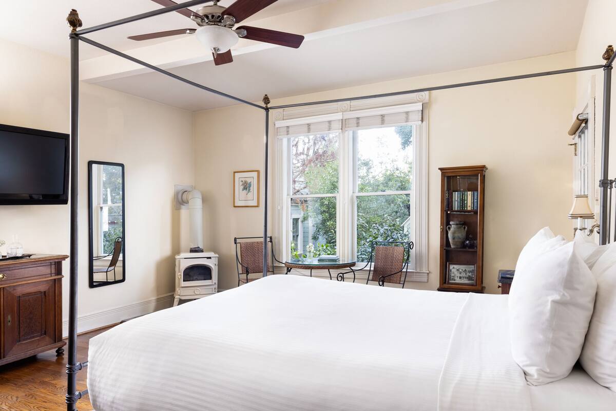 Quarto com cama de dossel, ventilador de teto, televisão, lareira e janela com vista para vegetação.
