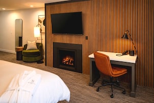 Quarto de hotel com cama, escrivaninha, cadeira, televisão, lareira, abajur e espelho.