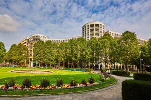 Um edifício moderno com um elemento circular central, rodeado por um jardim bem cuidado com um percurso circular e bancos.