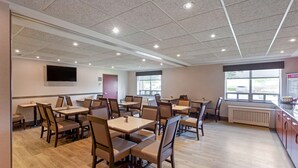 Uma área de jantar moderna com várias mesas e cadeiras, uma TV de tela plana na parede e grandes janelas que proporcionam luz natural.