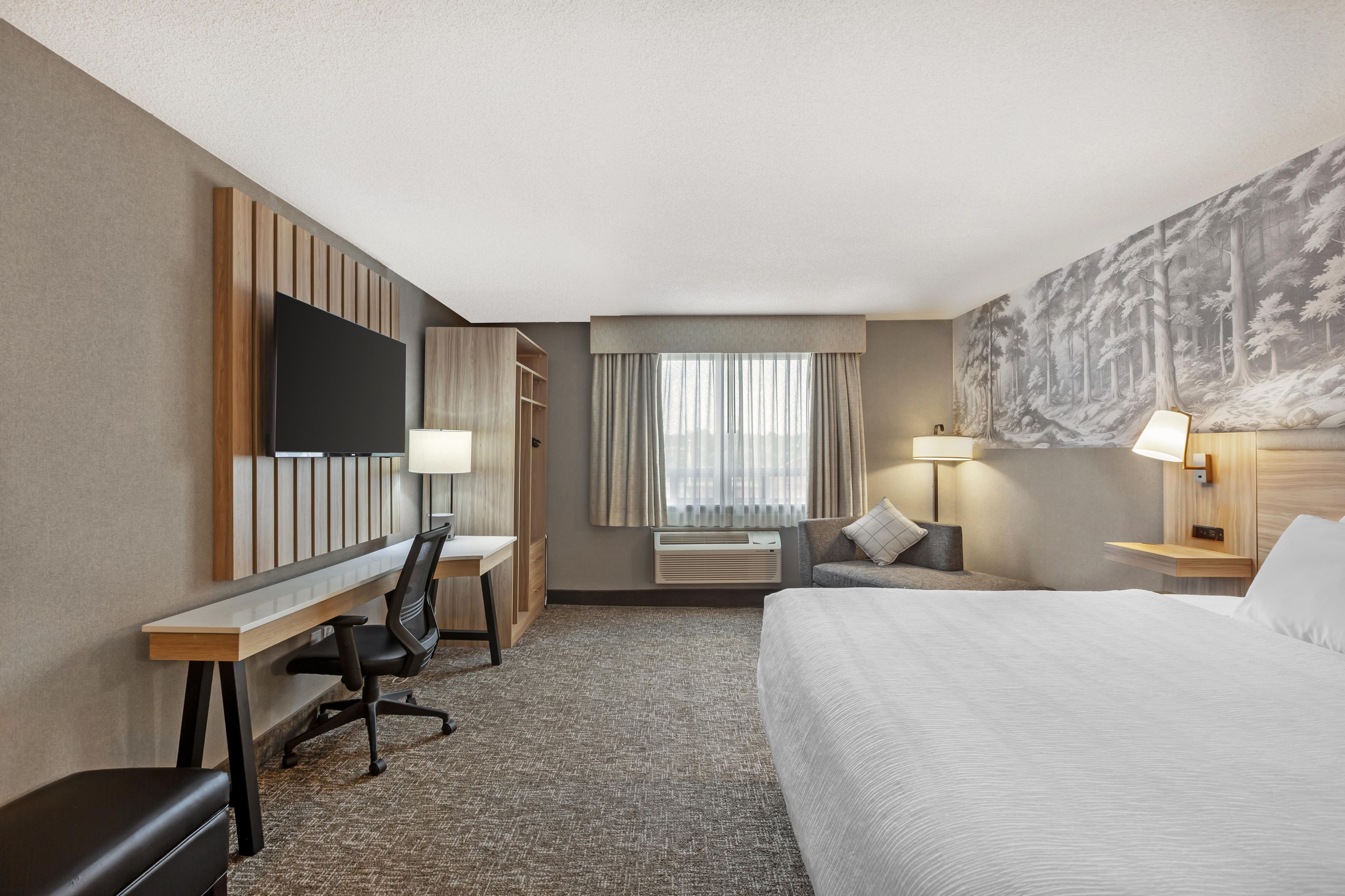 Quarto de hotel com cama, escrivaninha, cadeira, televisão e uma janela grande com cortinas.