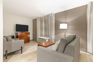 Quarto de hotel moderno com sofá cinzento, mesa de centro de madeira, televisão de tela plana, cômoda de madeira e uma grande janela com cortinas.