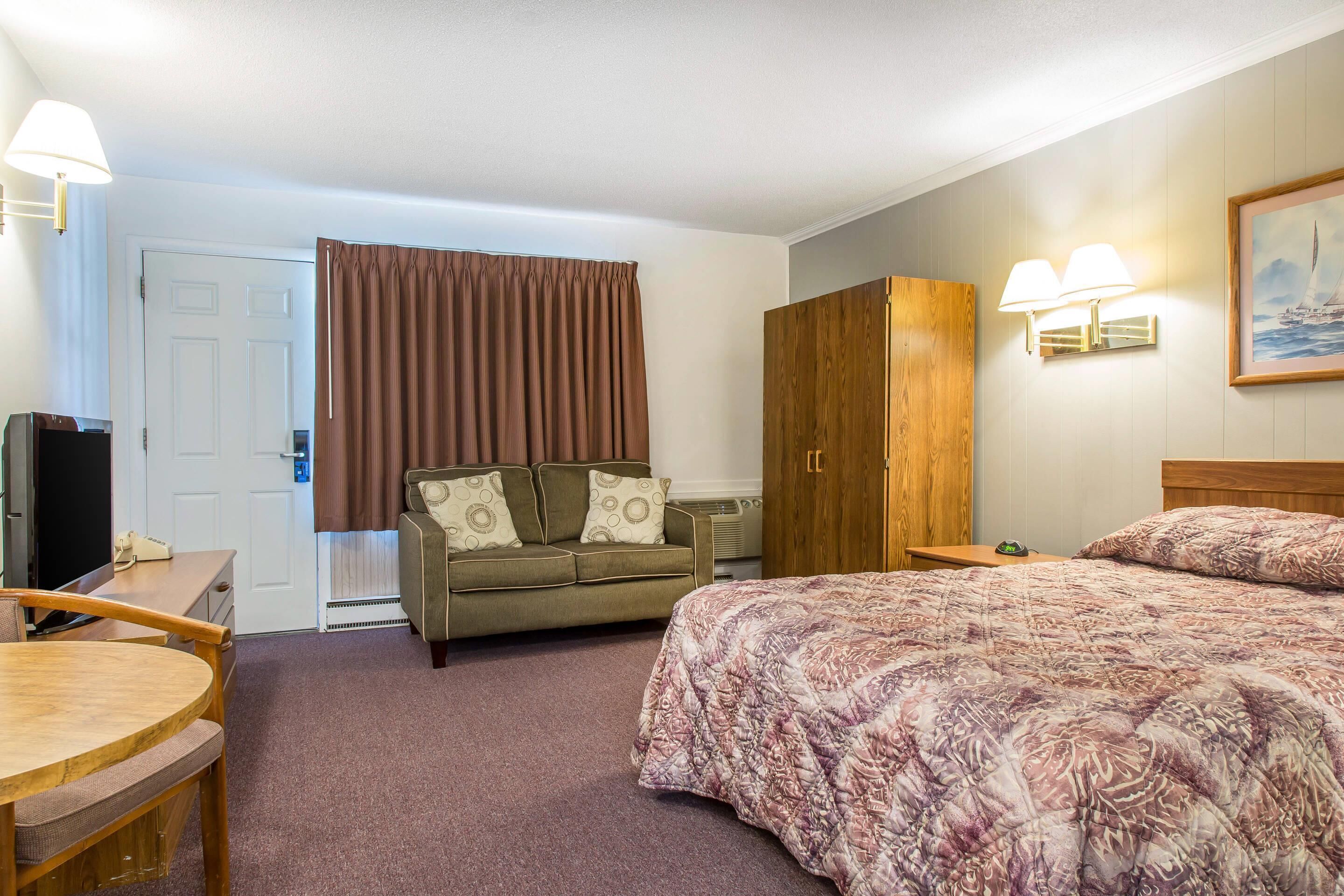Quarto de hotel com cama, sofá, guarda-roupa de madeira, escrivaninha com cadeira e televisão.