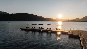 Um cais com espreguiçadeiras e guarda-sóis estendendo-se sobre águas calmas, com montanhas ao fundo e o sol a pôr-se.