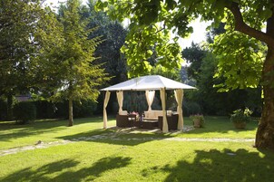 Um gazebo com teto branco e cortinas bege, cercado por vegetação exuberante e um caminho de pedra.