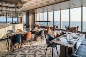 Restaurante com vista para o mar, com área de bar com garrafas e mesas com cadeiras dispostas para refeições.