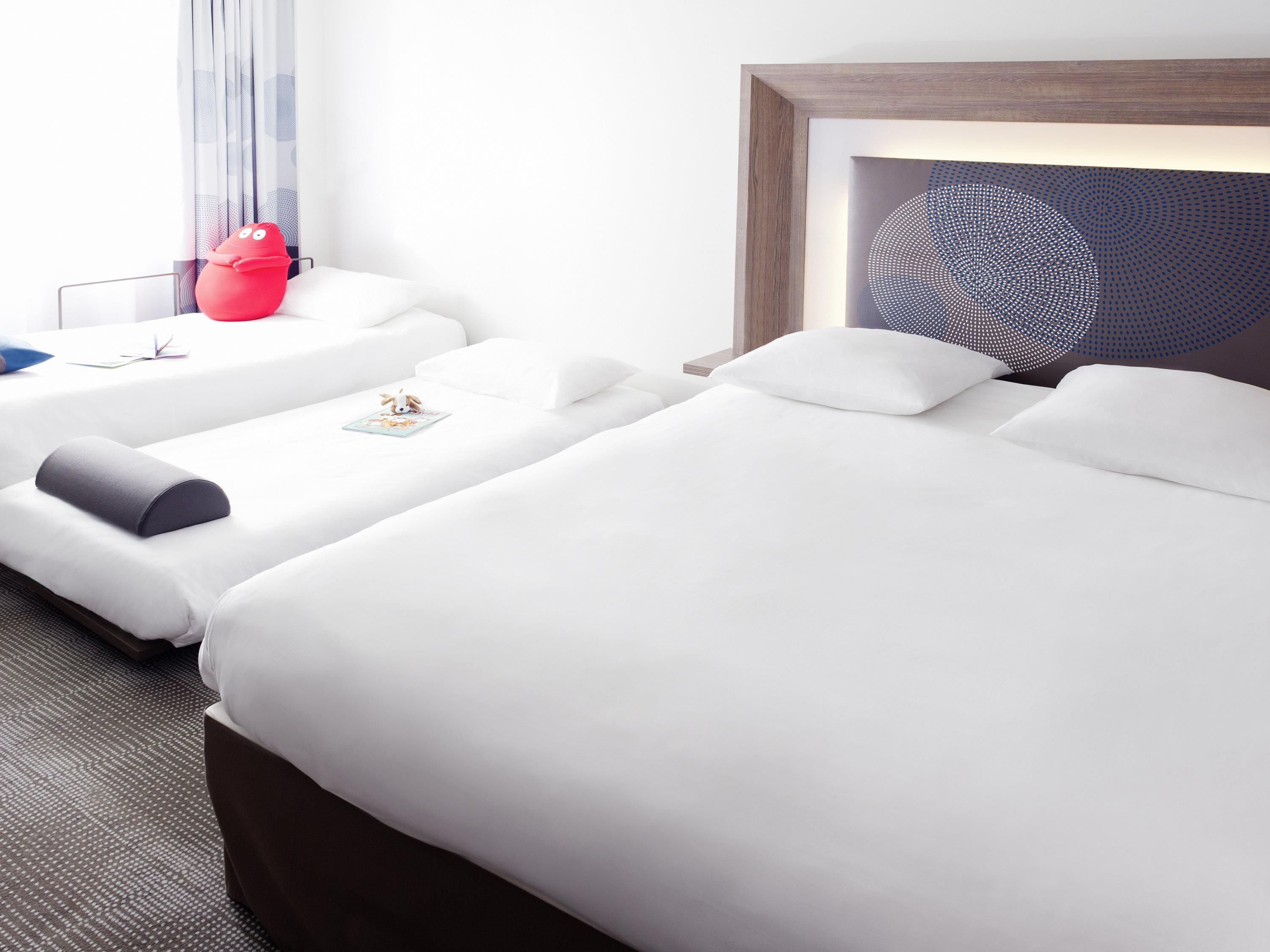 Quarto de hotel com duas camas, um brinquedo vermelho, uma obra de arte emoldurada e uma janela com cortinas.