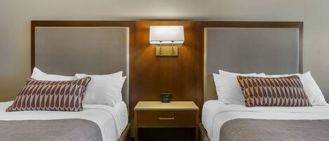 Quarto de hotel com duas camas, cada uma com almofada estampada, mesa de cabeceira com relógio e luminária fixa na parede.