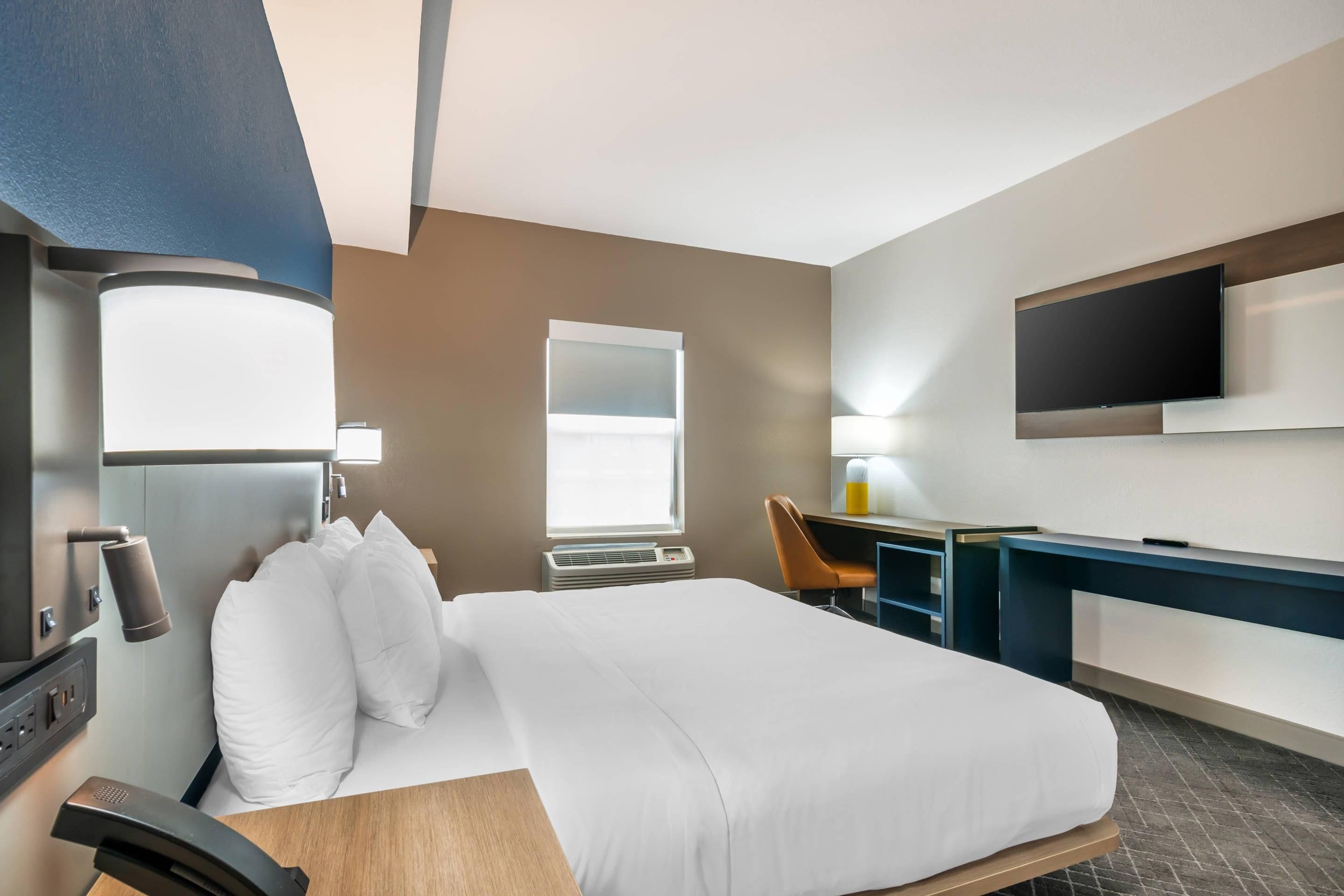Quarto de hotel com cama, escrivaninha, cadeira, televisão e janela.