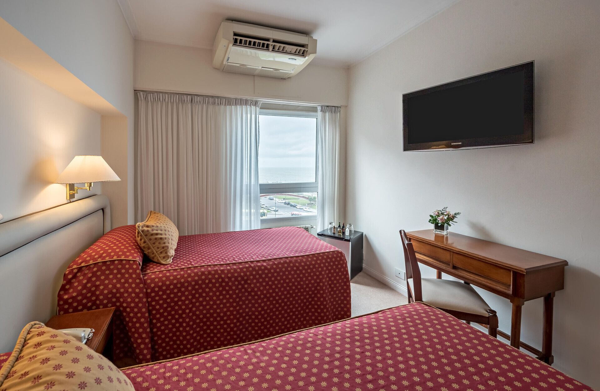 Quarto de hotel com duas camas, uma mesa, uma televisão e uma janela com cortinas.