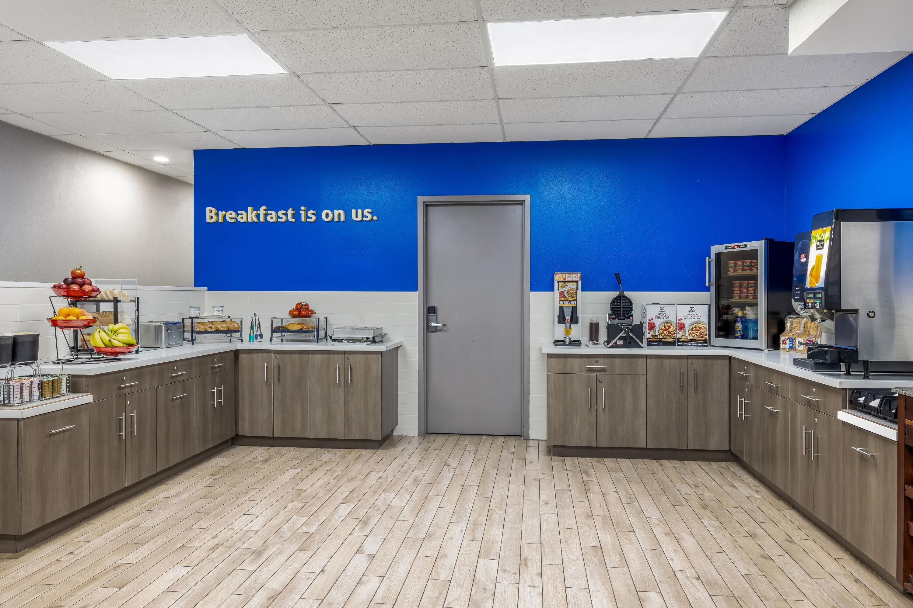 Espaço de café da manhã com parede azul, um aviso indicando 'Café da manhã por nossa conta' e diversas opções de alimentos e bebidas.