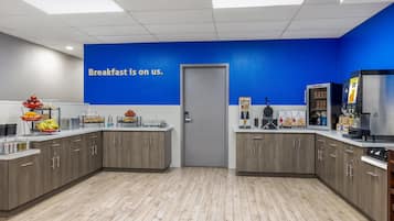 Espaço de café da manhã com parede azul, um aviso indicando 'Café da manhã por nossa conta' e diversas opções de alimentos e bebidas.