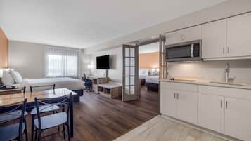 Quarto de hotel com cama, escrivaninha, cadeira, televisão, micro-ondas e cozinha compacta.