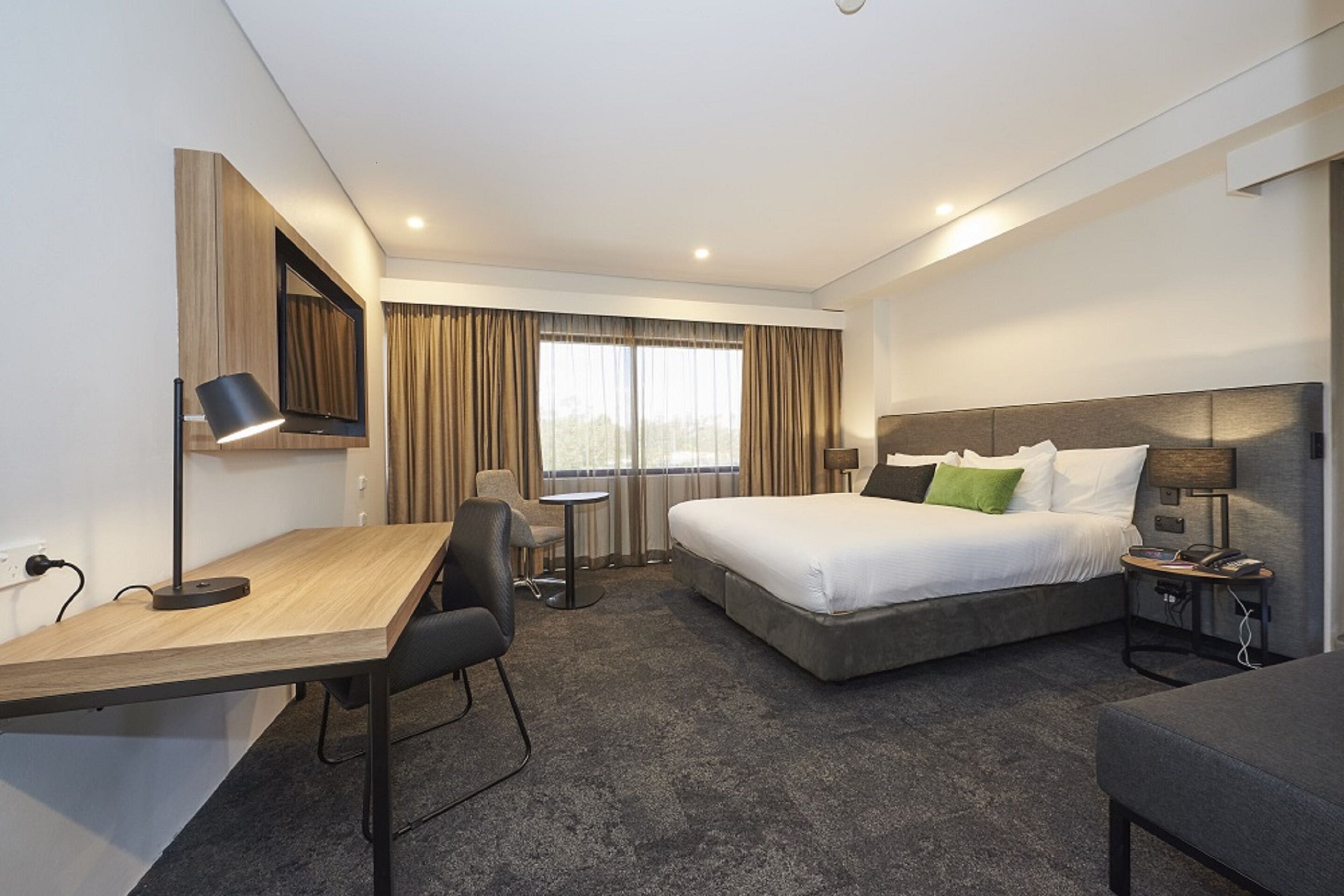 Quarto de hotel moderno com uma cama grande, uma escrivaninha, uma cadeira, uma mesinha e uma televisão fixada na parede.