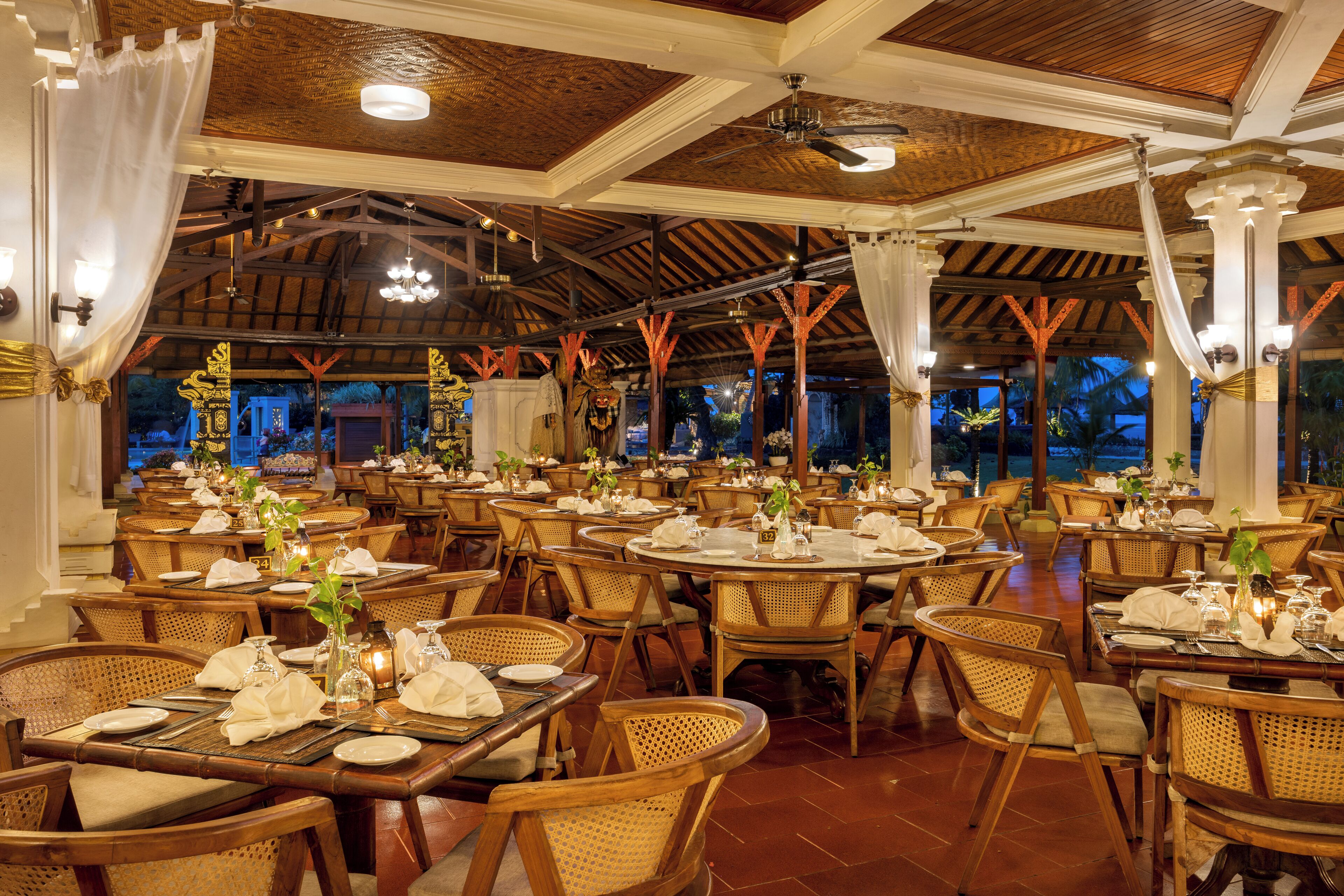 Um restaurante com mesas redondas, toalhas brancas, cadeiras de vime e centros de mesa com velas.