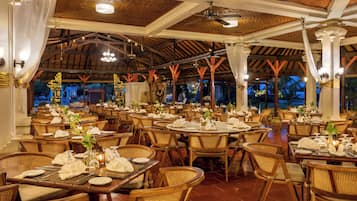 Um restaurante com mesas redondas, toalhas brancas, cadeiras de vime e centros de mesa com velas.