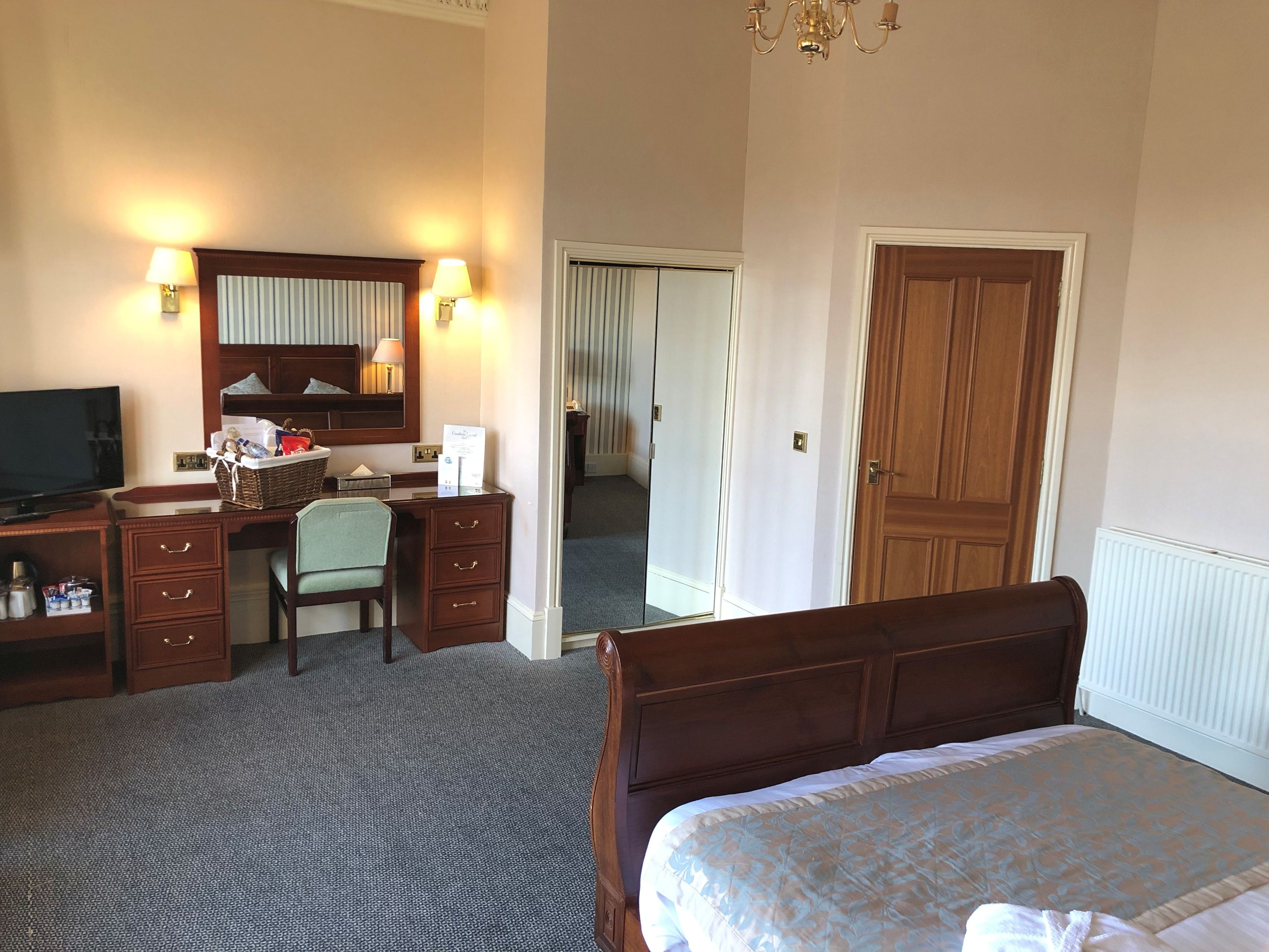 Quarto de hotel com cama, escrivaninha, computador, cadeira, televisão e espelho.