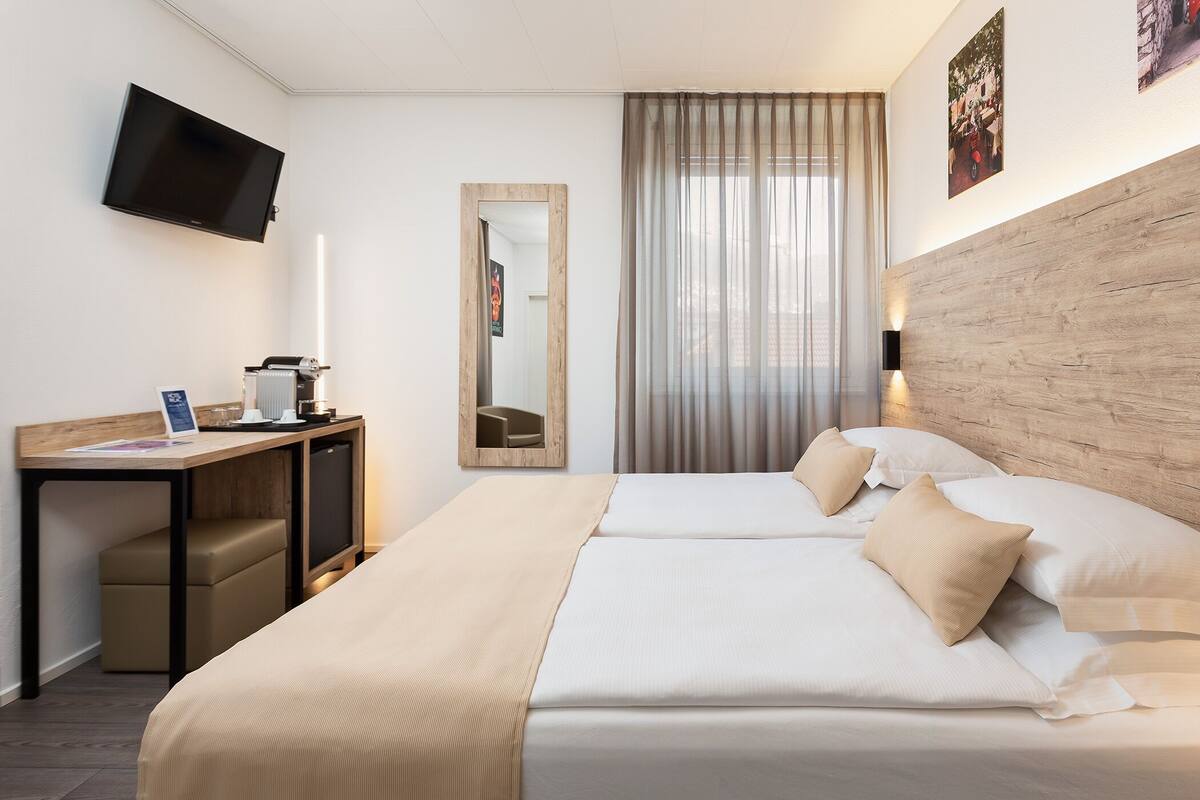 Quarto de hotel com cama, escrivaninha com computador, televisão e espelho.