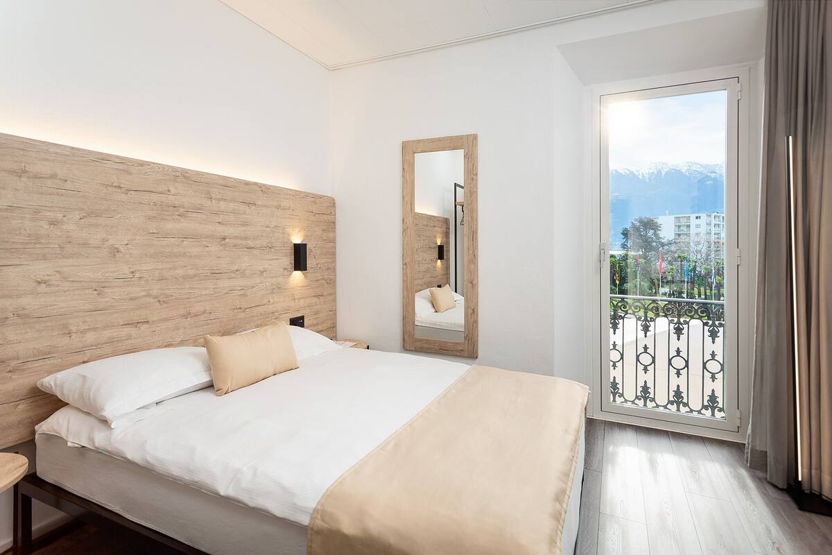 Quarto de hotel com cabeceira de madeira, cama grande com roupa de cama branca, espelho e varanda com vista para montanhas.