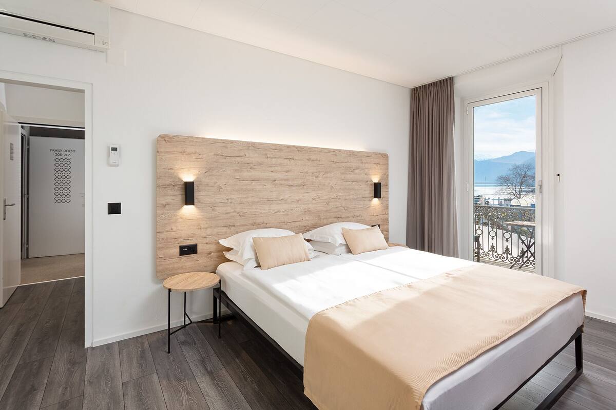 Quarto de hotel moderno com uma cama grande, cabeceira de madeira, luminárias laterais e vista da cidade através de uma varanda.