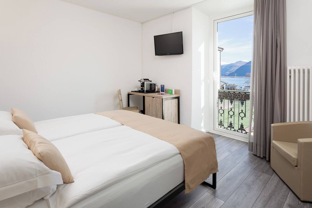 Quarto de hotel com cama, secretária com cafeteira, televisão e varanda com vista para montanhas e lago.