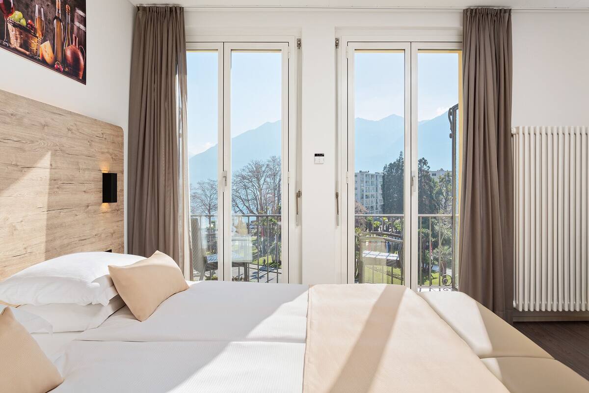 Quarto de hotel com cama, cabeceira de madeira, uma janela grande com cortinas e vista para montanhas.