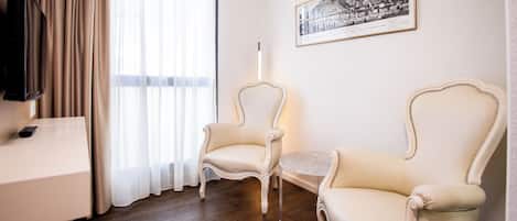 Quarto de hotel com chão de madeira, uma cama, televisão, duas poltronas brancas, uma mesinha e um quadro emoldurado na parede.