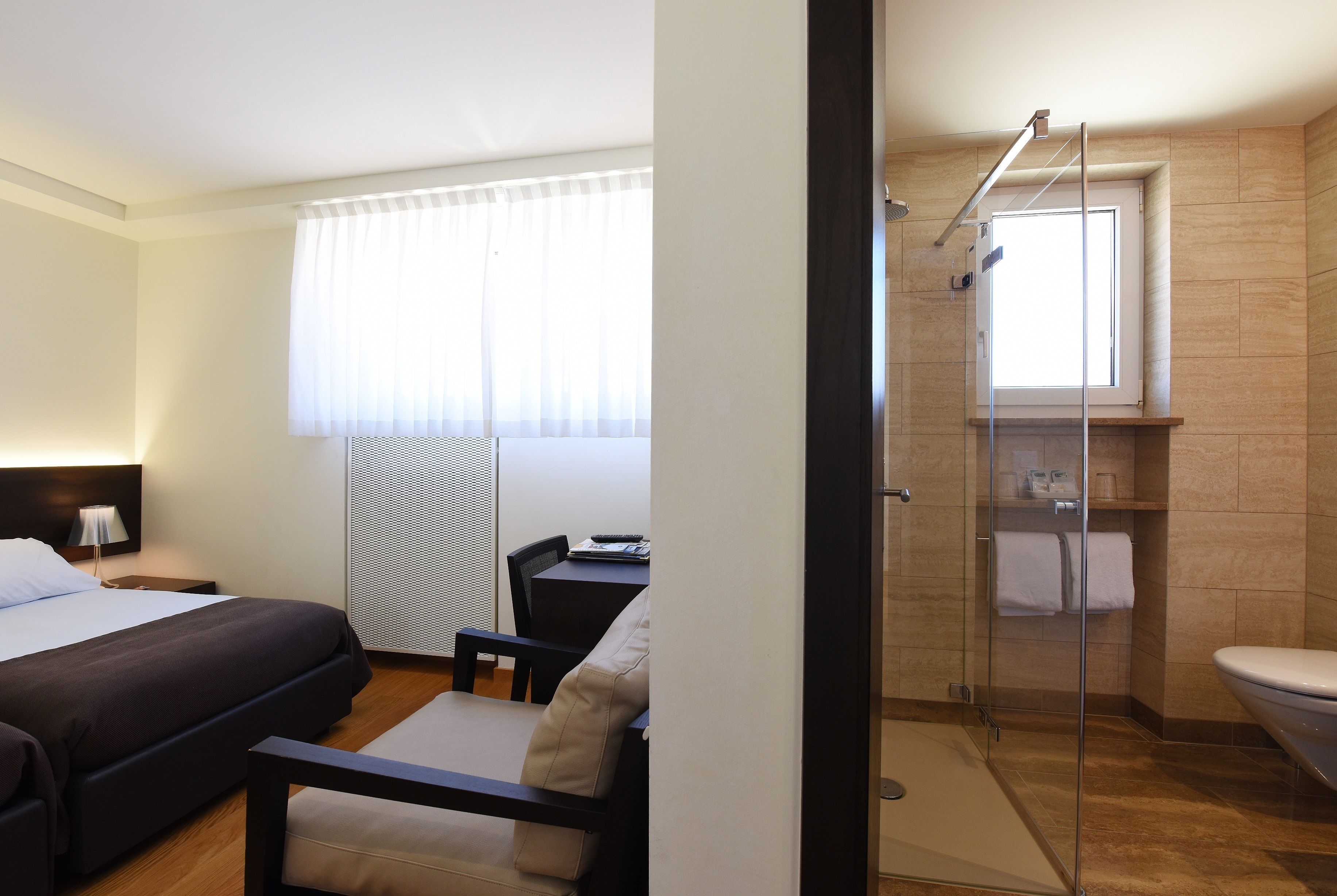 Quarto de hotel com cama, escrivaninha, cadeira, chuveiro e banheiro.