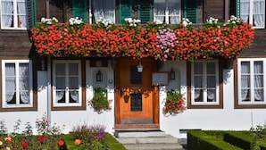 Um edifício tradicional com porta de madeira, paredes brancas e portadas verdes. Há caixas de flores com flores vermelhas e cor-de-rosa sobre a entrada.