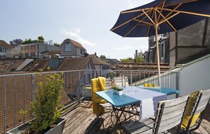 Terraço no telhado com mesa e cadeiras, um guarda-sol azul e vista para edifícios ao redor.