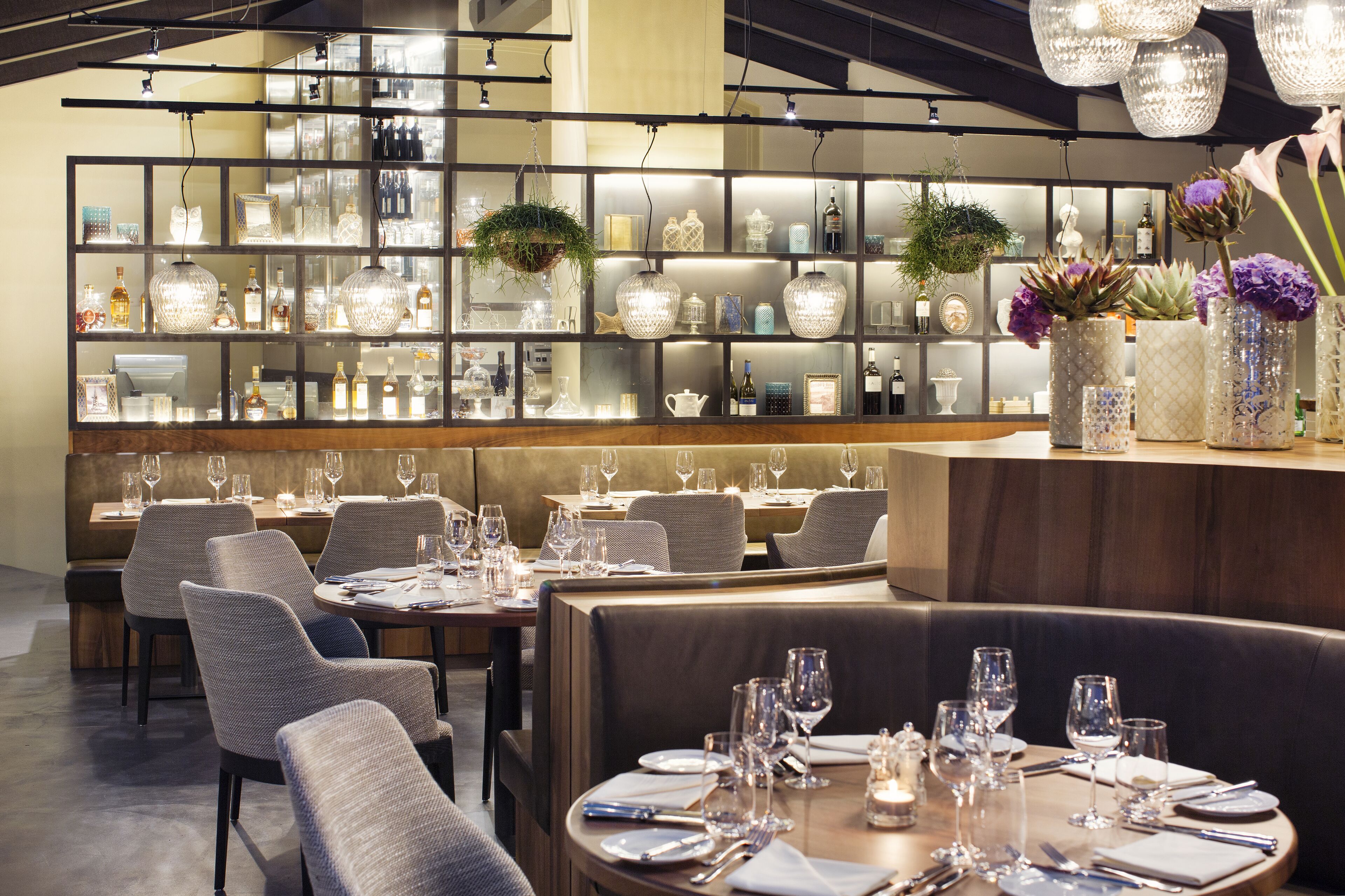 Restaurante com diversas mesas preparadas para os clientes, área de bar e exposição de diversos copos e objetos decorativos nas prateleiras.