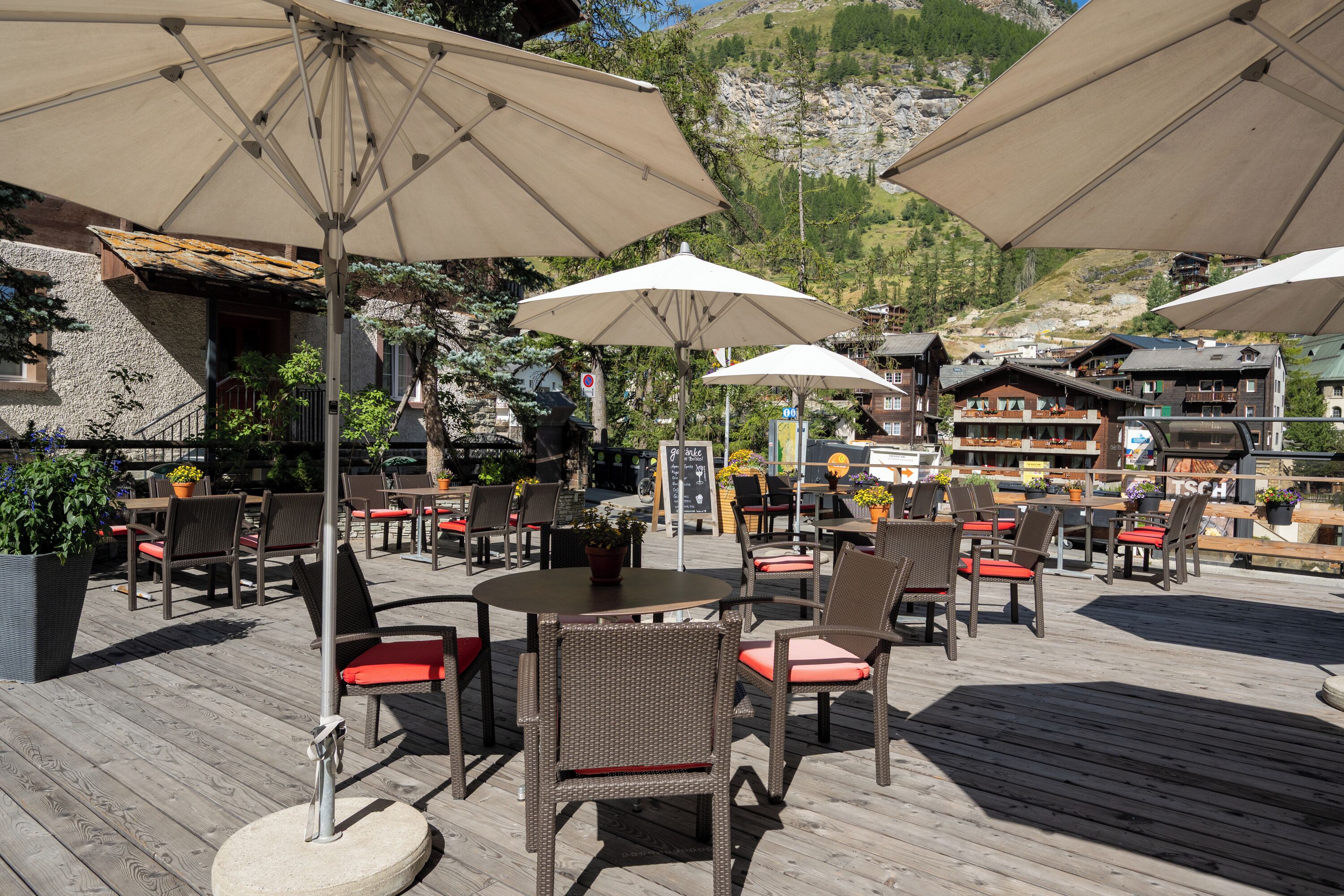 Um terraço ao ar livre com cadeiras e mesas de vime, guarda-sóis e vista para montanhas e edifícios.