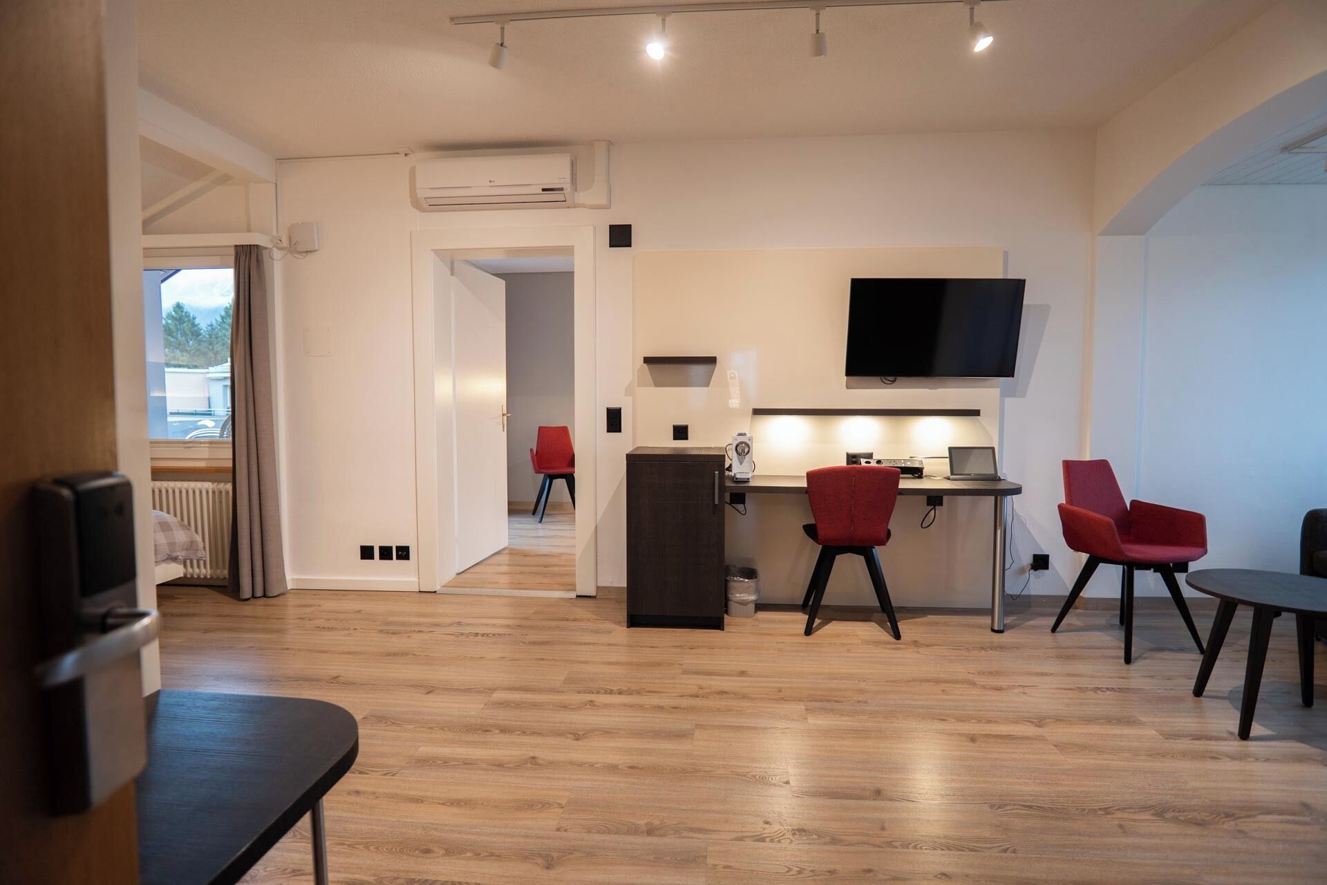 Quarto de hotel moderno com piso de madeira, uma escrivaninha com um laptop, televisão de tela plana e cadeiras vermelhas.