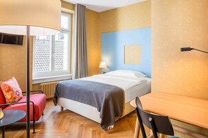 Quarto de hotel com cama, escrivaninha, cadeira, abajur, televisão e janela com cortinas.