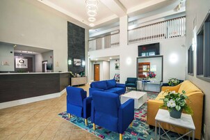 Um saguão de hotel com balcão de recepção, assentos em azul e amarelo, uma mesa de centro e uma planta em vaso.