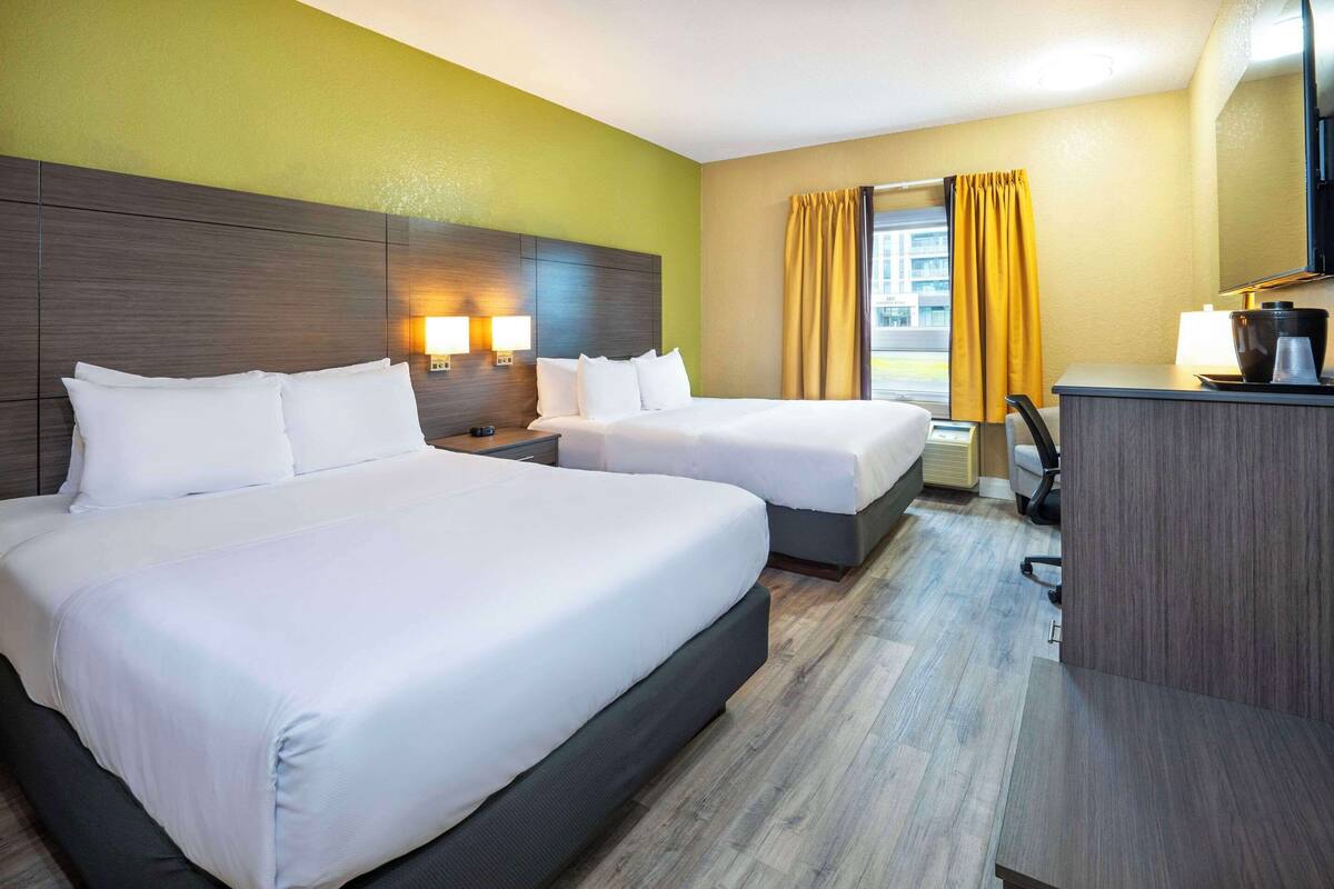 Quarto de hotel com duas camas, uma escrivaninha, uma cadeira e uma janela com cortinas.