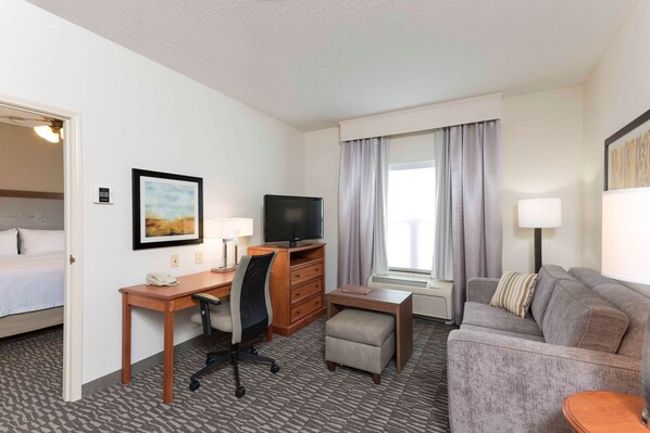 Quarto de hotel com uma cama, uma escrivaninha com um computador, uma televisão, um sofá e uma janela com cortinas.