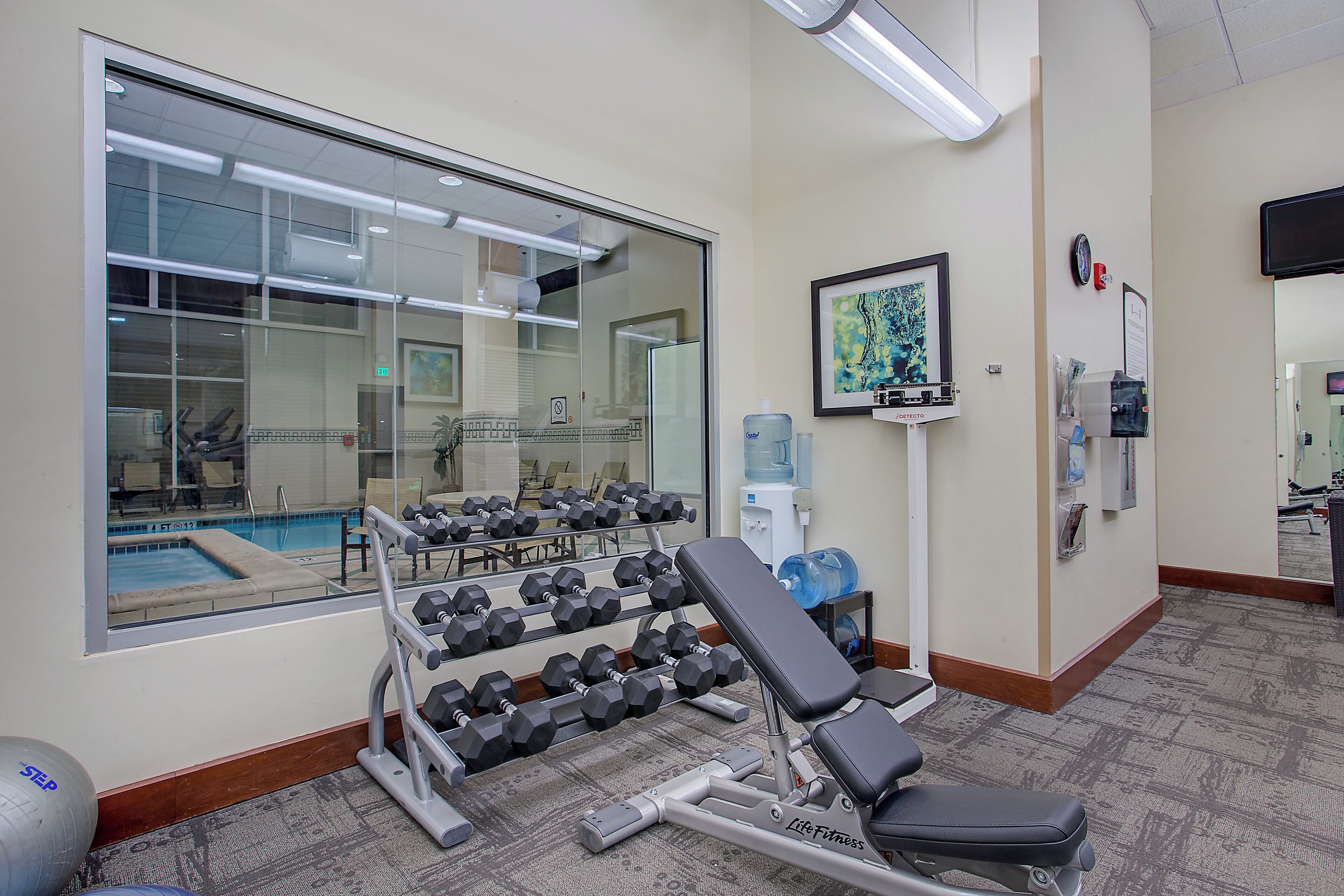 Academia com equipamentos de ginástica, bebedouro e piscina visíveis através da parede de vidro.