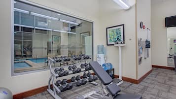 Academia com equipamentos de ginástica, bebedouro e piscina visíveis através da parede de vidro.