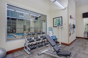 Academia com equipamentos de ginástica, bebedouro e piscina visíveis através da parede de vidro.
