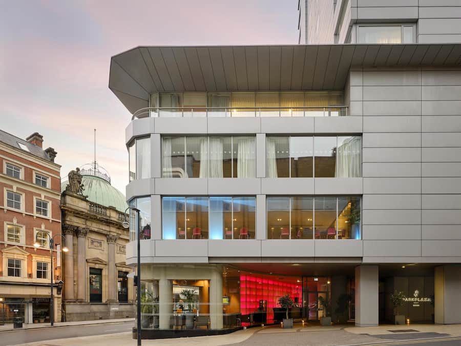 Um hotel moderno com fachada de vidro, entrada iluminada em vermelho e um edifício clássico ao lado.
